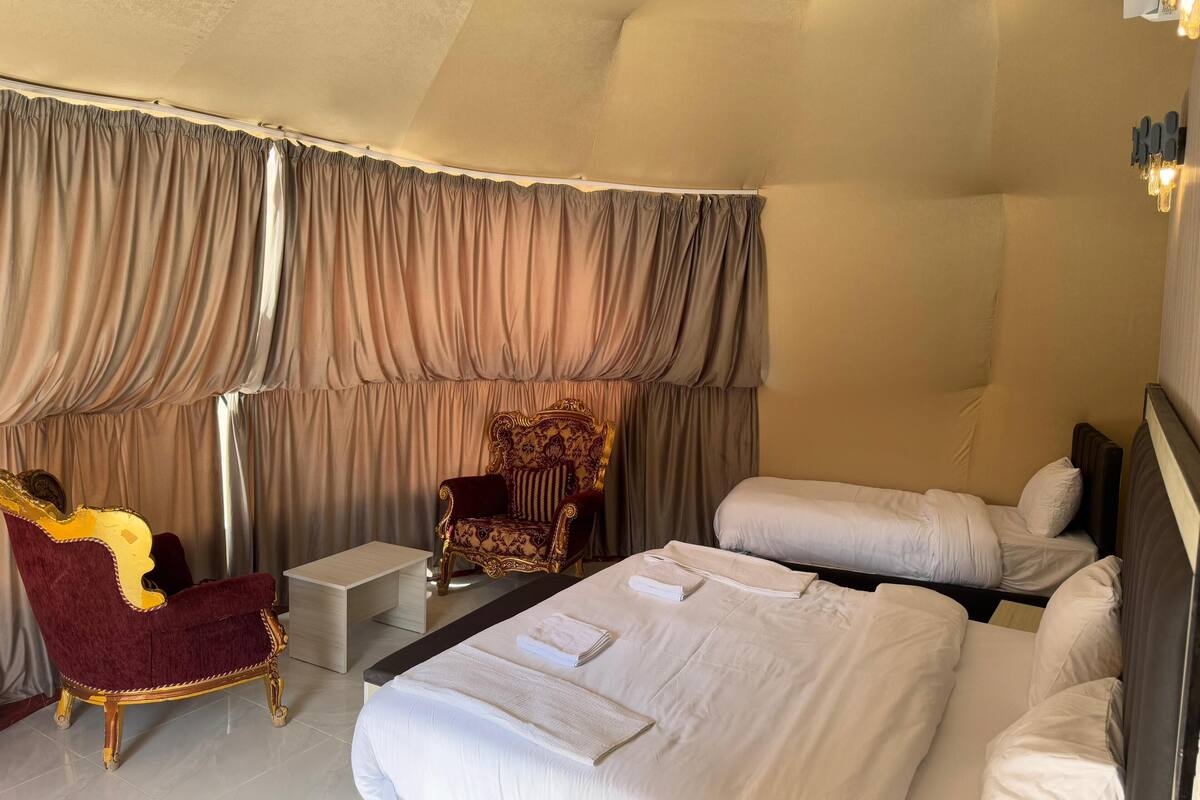 Quarto de hotel com uma cama grande, uma cama menor, uma toucador com espelho, uma cadeira e uma janela grande com cortinas.