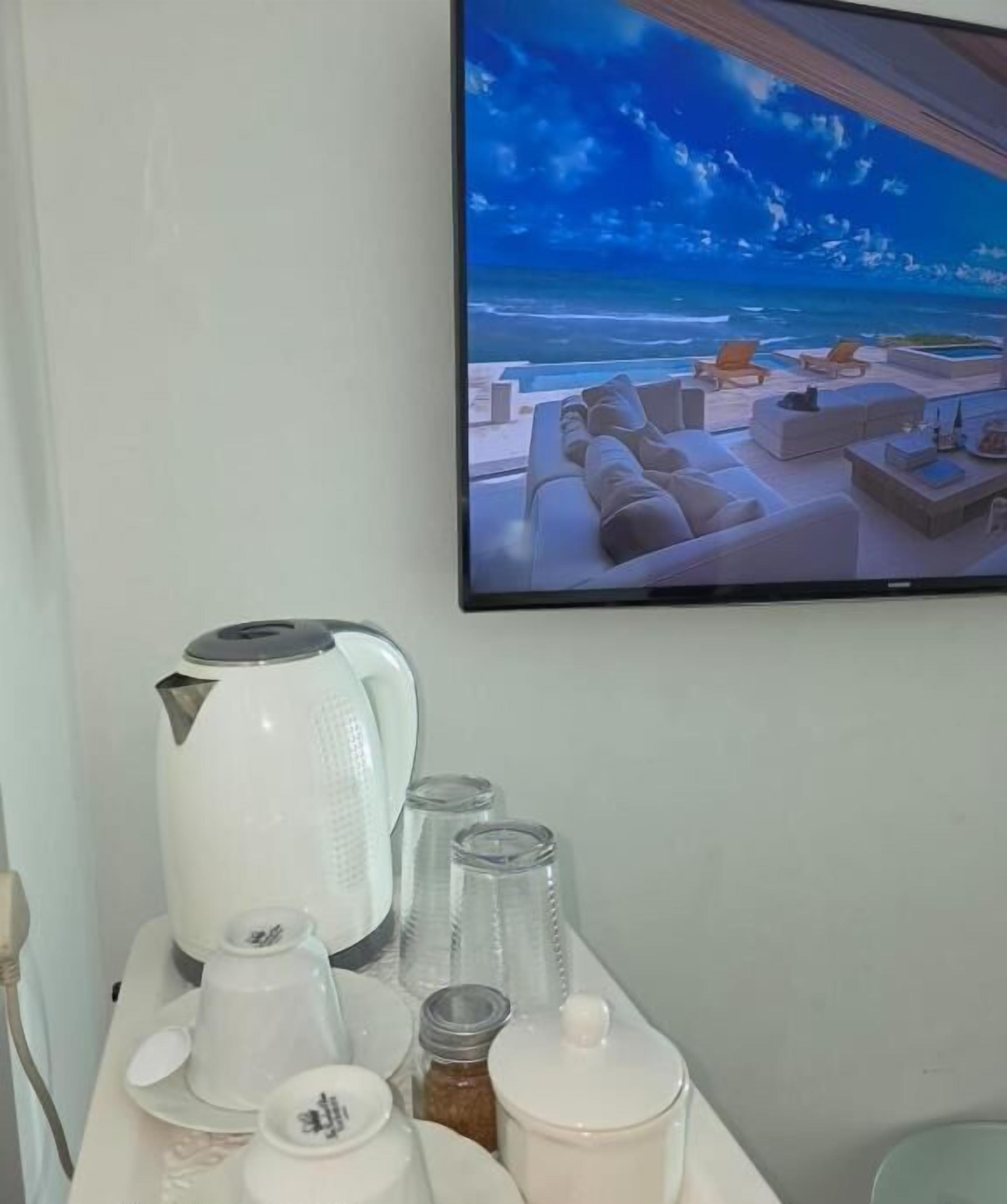Quarto com uma televisão de tela grande exibindo uma cena de praia, um espaço para café com chaleira, xícaras e potes, e uma parede branca.