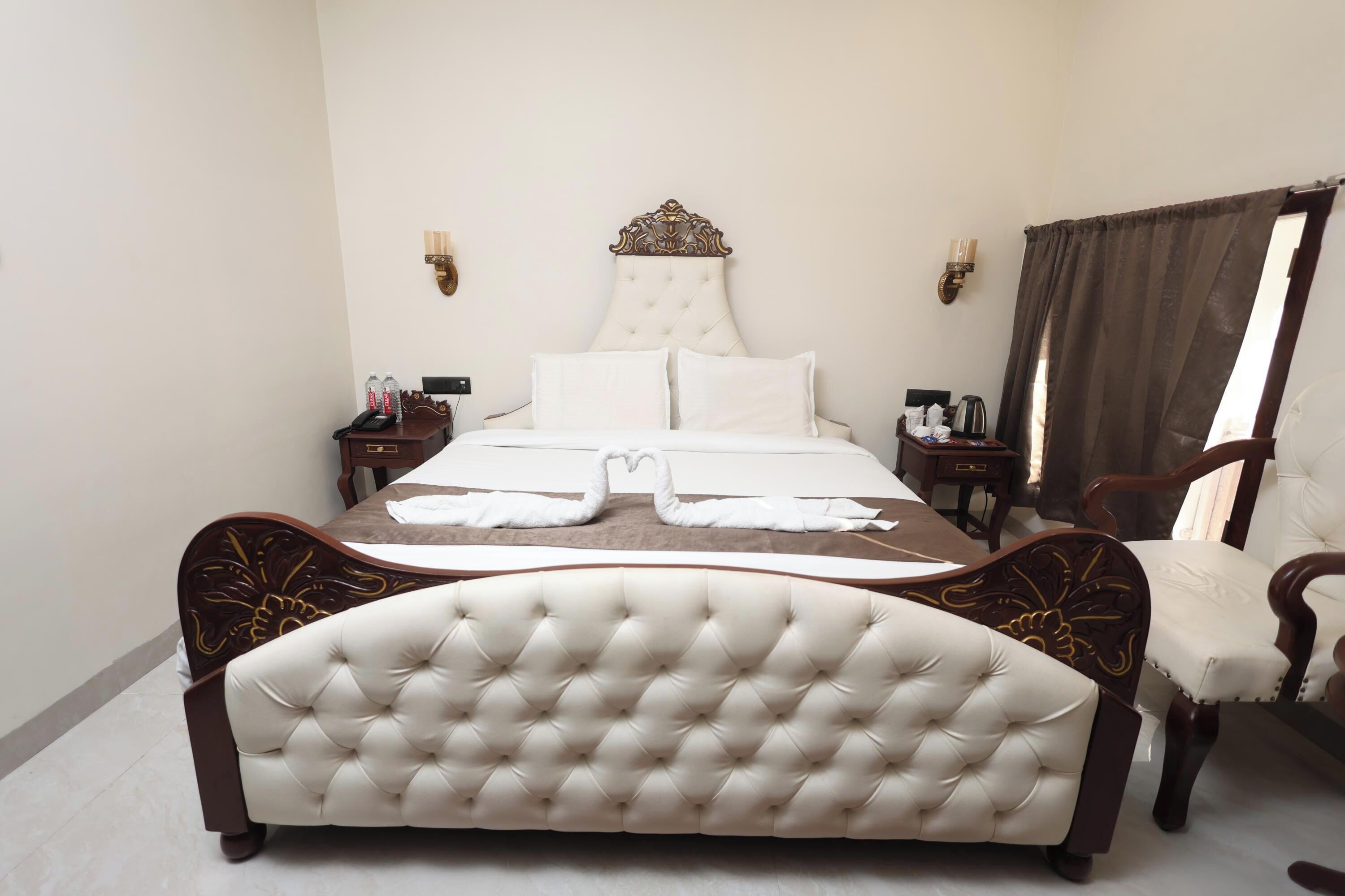 Uma cama bem arrumada com lençóis brancos e almofadas decorativas, uma cabeceira estofada, uma mesa de cabeceira de madeira com uma luminária e uma cadeira com assento acolchoado.