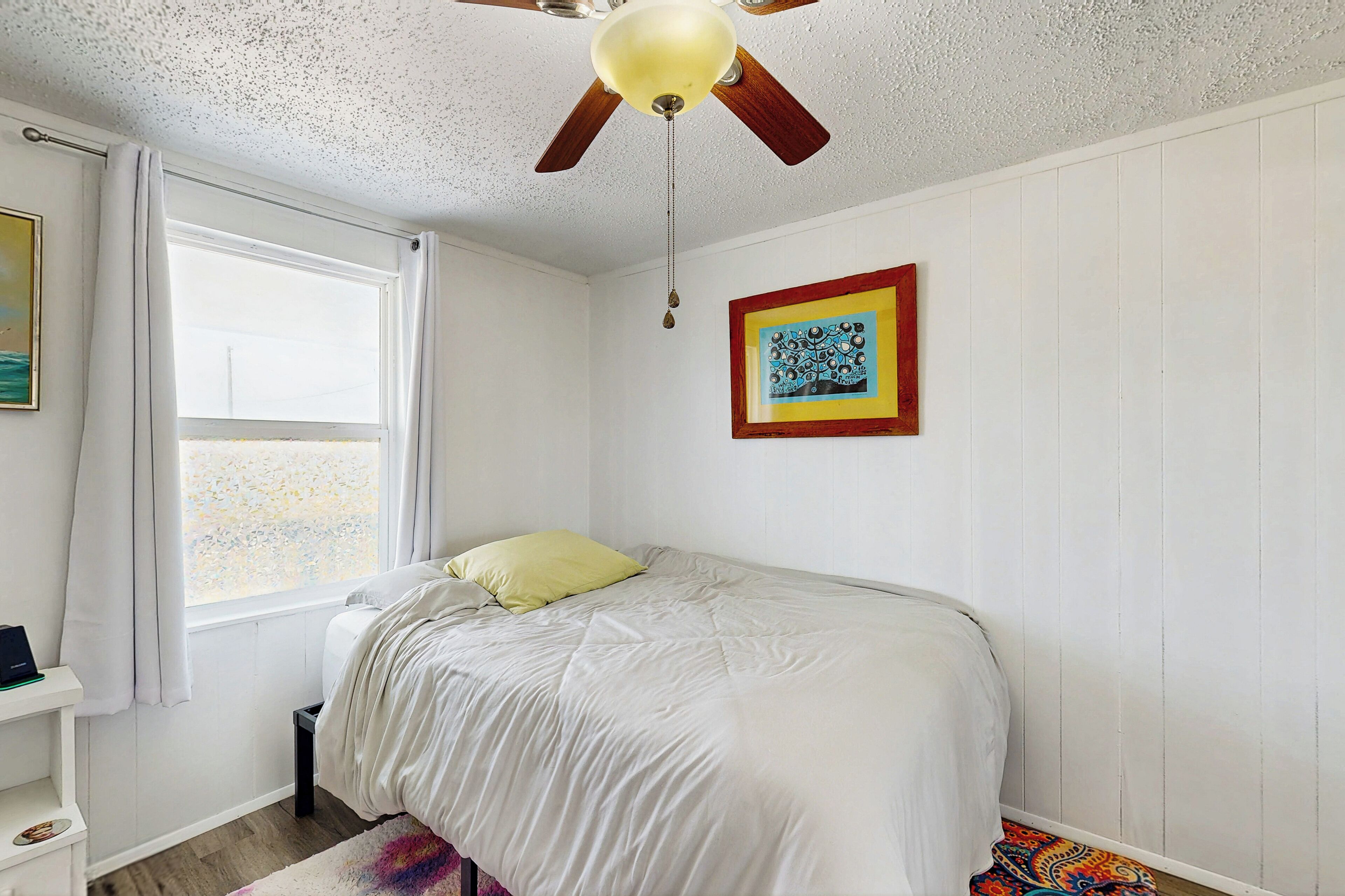 Quarto com cama, ventilador de teto, janela com cortinas, quadro pendurado na parede e pequena mesa com luminária.