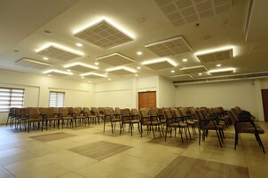 Uma sala de conferências com fileiras de cadeiras, luminárias no teto e uma porta de madeira.