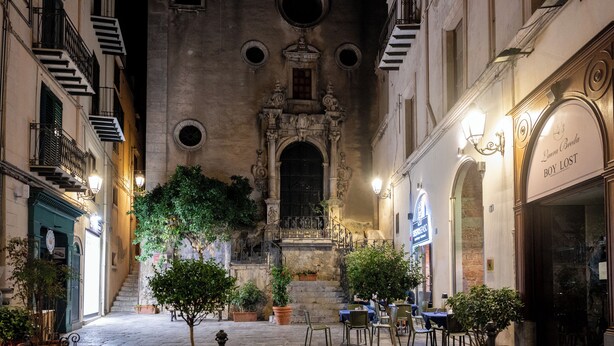 Um edifício histórico com uma entrada central em arco, ladeada por varandas e plantas em vasos.