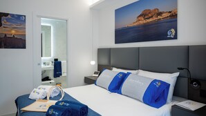 Quarto de hotel com cama, mesa de cabeceira, escrivaninha, cadeira, espelho e um quadro de uma paisagem costeira.