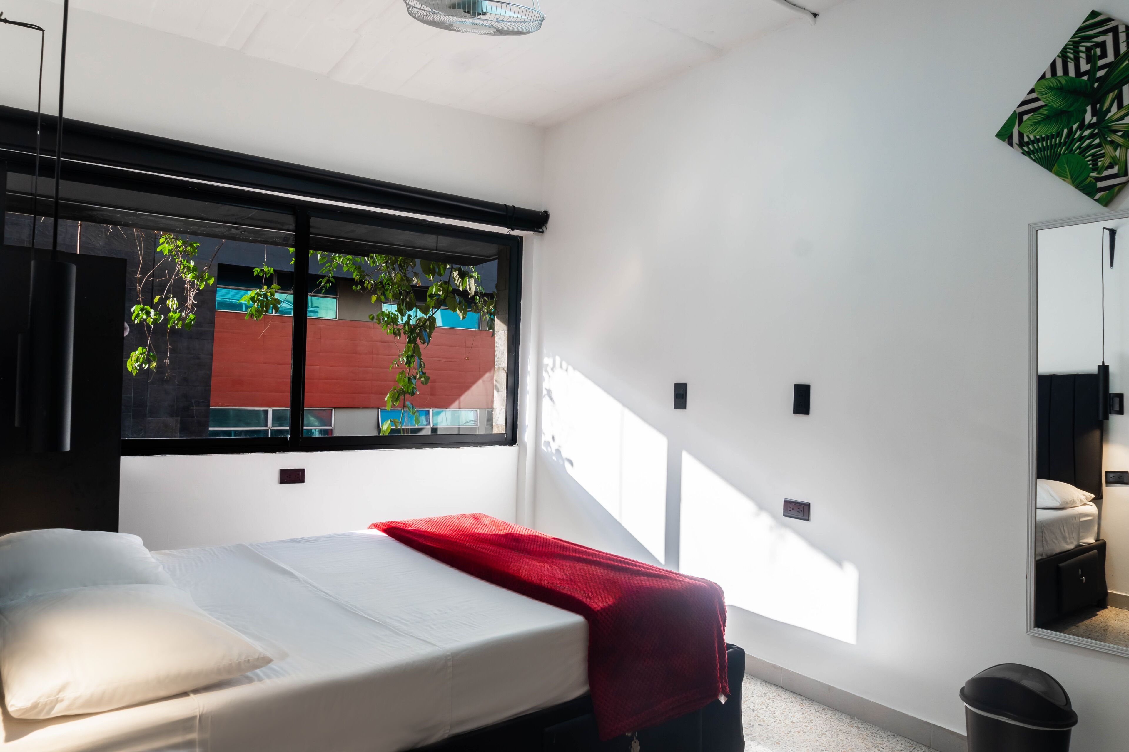 Quarto de hotel moderno com cama, janela com vista para edifícios, manta vermelha, lixeira preta e espelho fixado na parede.