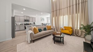 Sala de estar moderna com sofá, mesa de centro e poltrona amarela. Cozinha visível ao fundo, com armários brancos e micro-ondas.