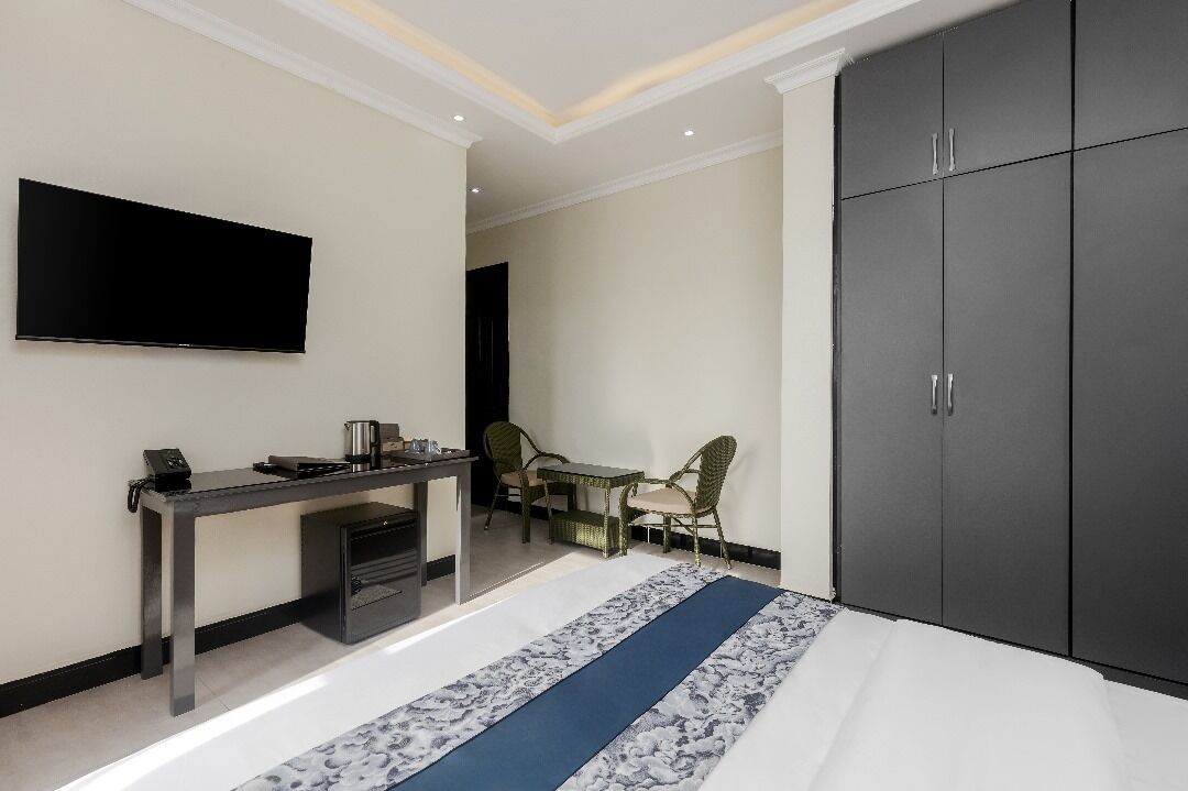 Quarto de hotel moderno com televisão de tela plana, escrivaninha com máquina de café, área de refeições com cadeiras e um guarda-roupa grande.