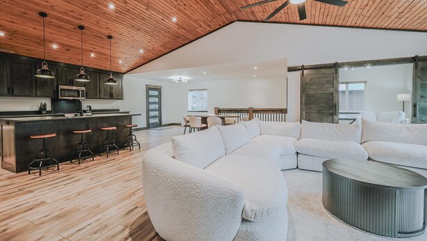 Uma sala de estar ampla com teto de madeira, um sofá seccional branco grande, uma mesa de centro redonda e uma área de cozinha com eletrodomésticos modernos.