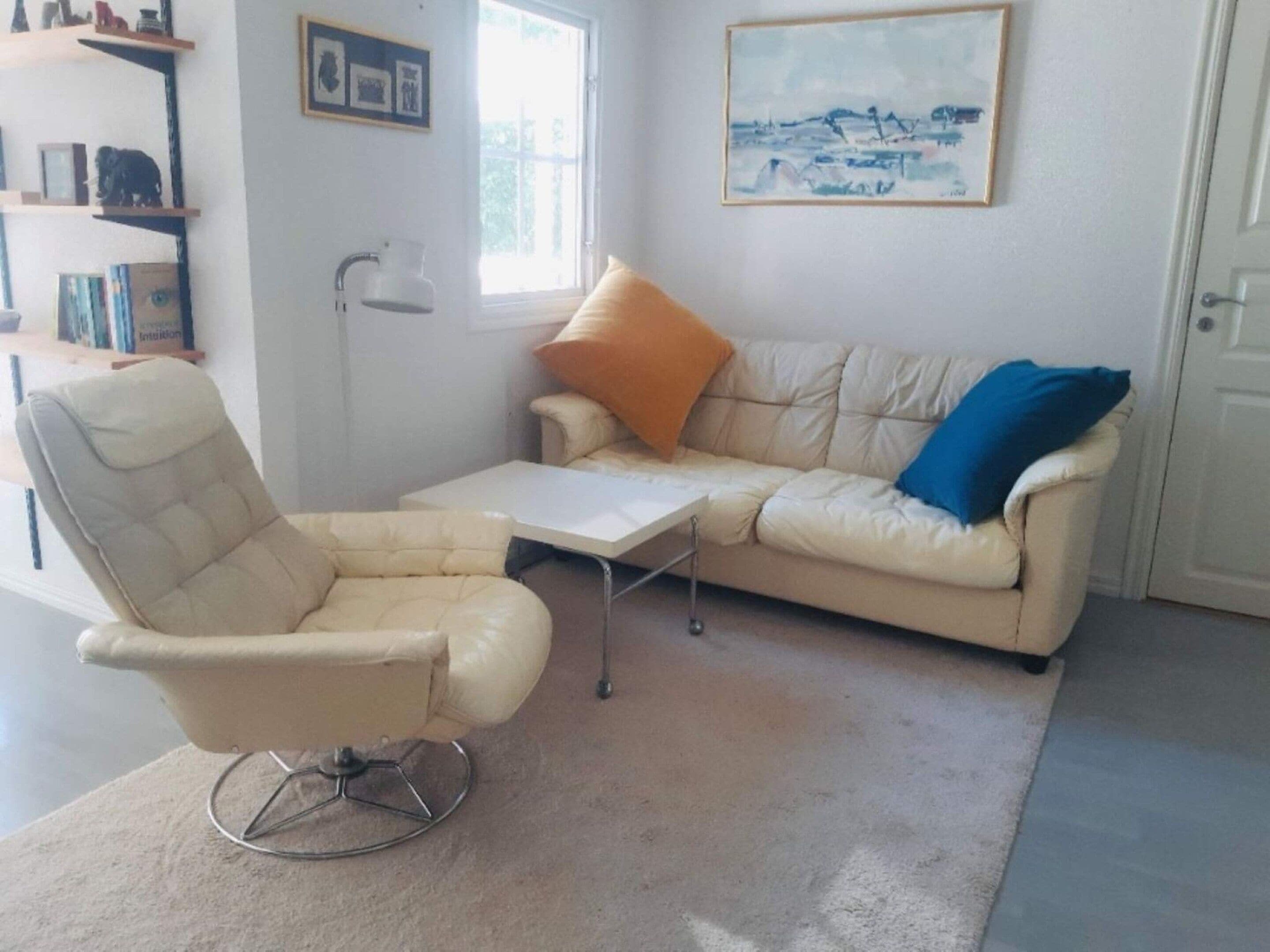 Uma sala de estar com sofá bege, poltrona branca, mesinha pequena branca e luminária de chão.