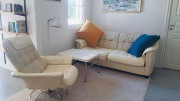 Uma sala de estar com sofá bege, poltrona branca, mesinha pequena branca e luminária de chão.