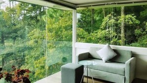 Quarto moderno com uma janela ampla que oferece vista para uma floresta. Há um sofá cinzento com uma mesinha e um vaso de planta.