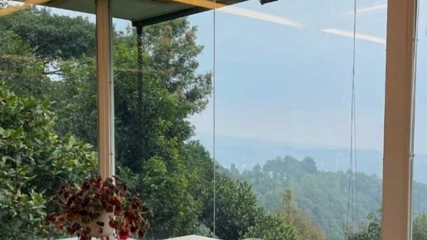 Uma sala de jantar moderna com uma mesa comprida, um vaso de flores e vista para uma paisagem arborizada através de grandes janelas.