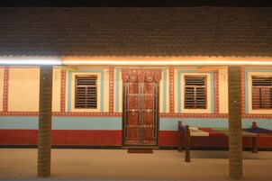 Um edifício tradicional com telhado em telhas, porta de madeira central com painéis decorativos e duas janelas com persianas.