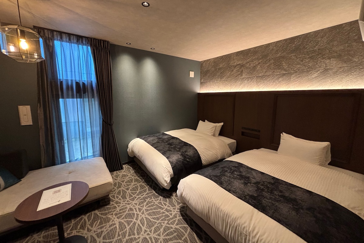 Quarto de hotel com duas camas, uma mesinha e janela com cortinas.