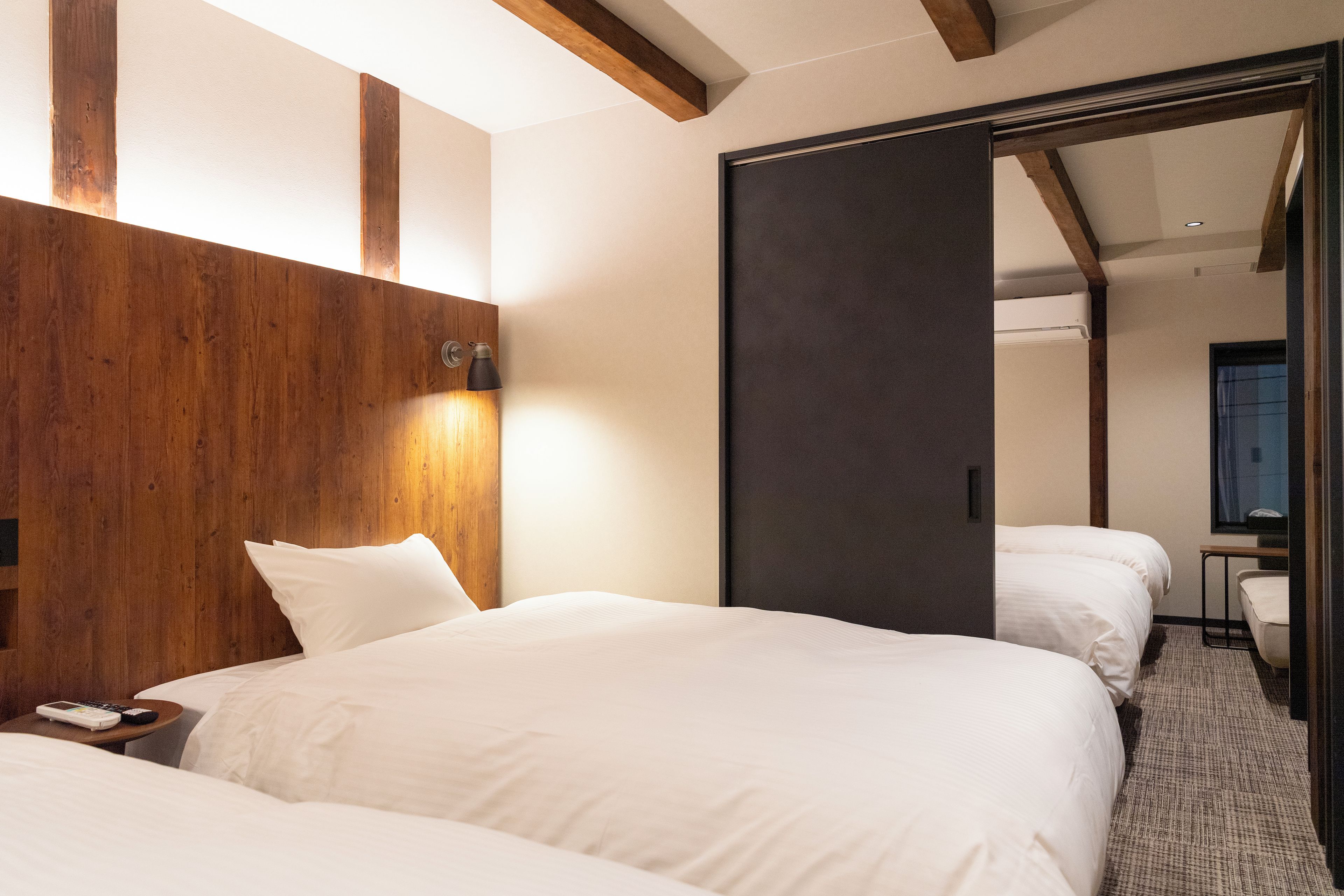 Quarto de hotel com duas camas, cabeceiras de madeira, uma porta escura e uma pequena mesa com um controle remoto.