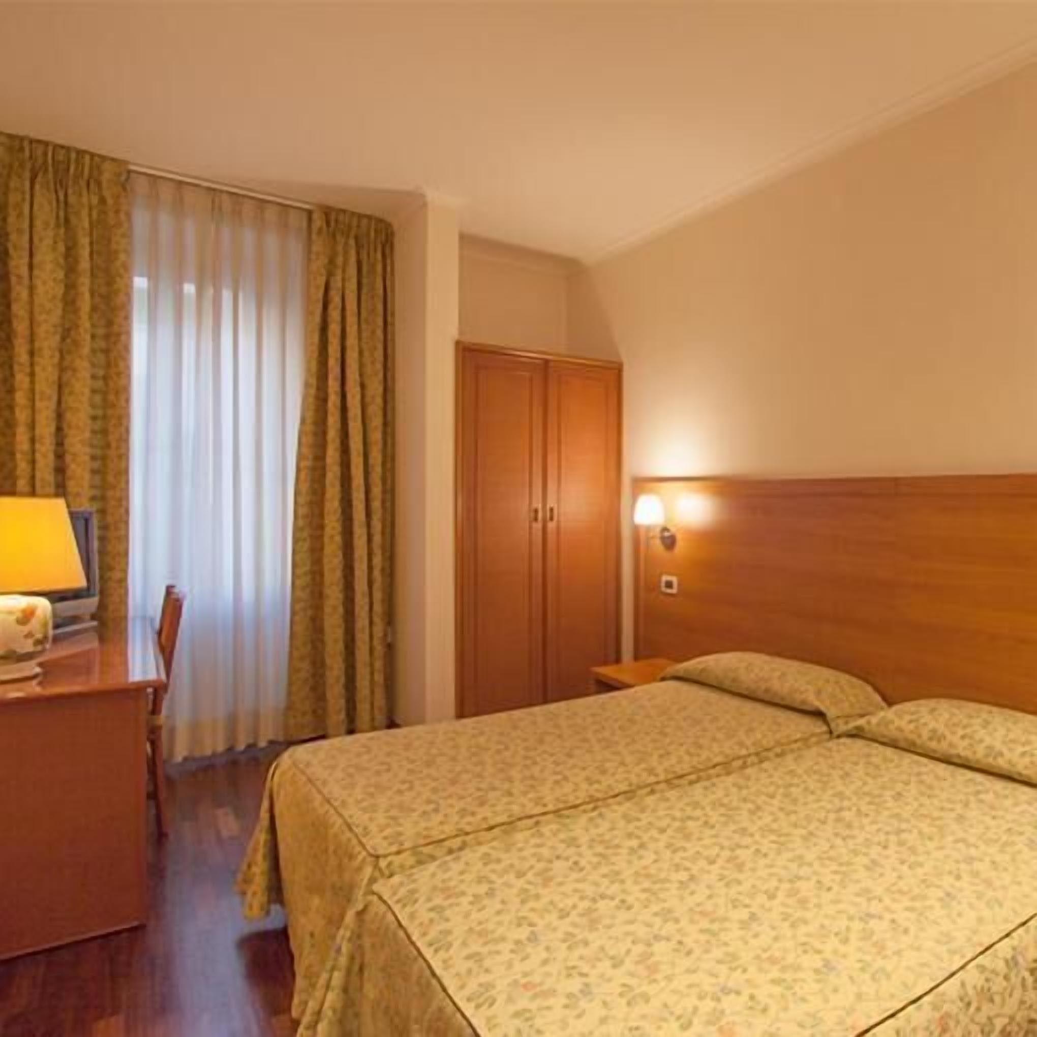 Quarto de hotel com cama, escrivaninha com abajur, guarda-roupa e janela com cortinas.