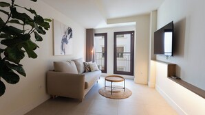 Uma sala de estar moderna com um sofá, uma mesa de centro redonda, uma TV fixada na parede e uma janela grande com vista para o exterior.