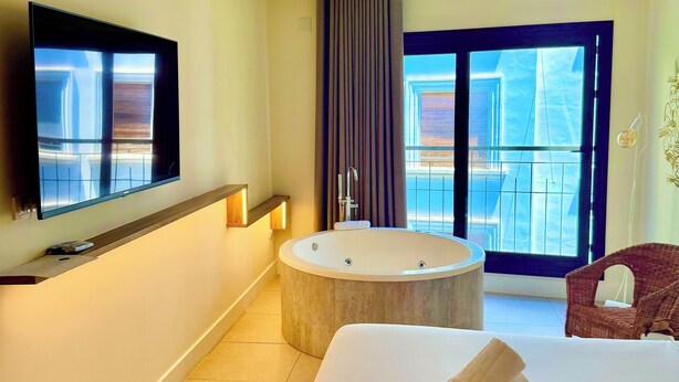 Quarto de hotel com uma cama grande, banheira redonda, televisão fixada na parede e uma varanda com vista.