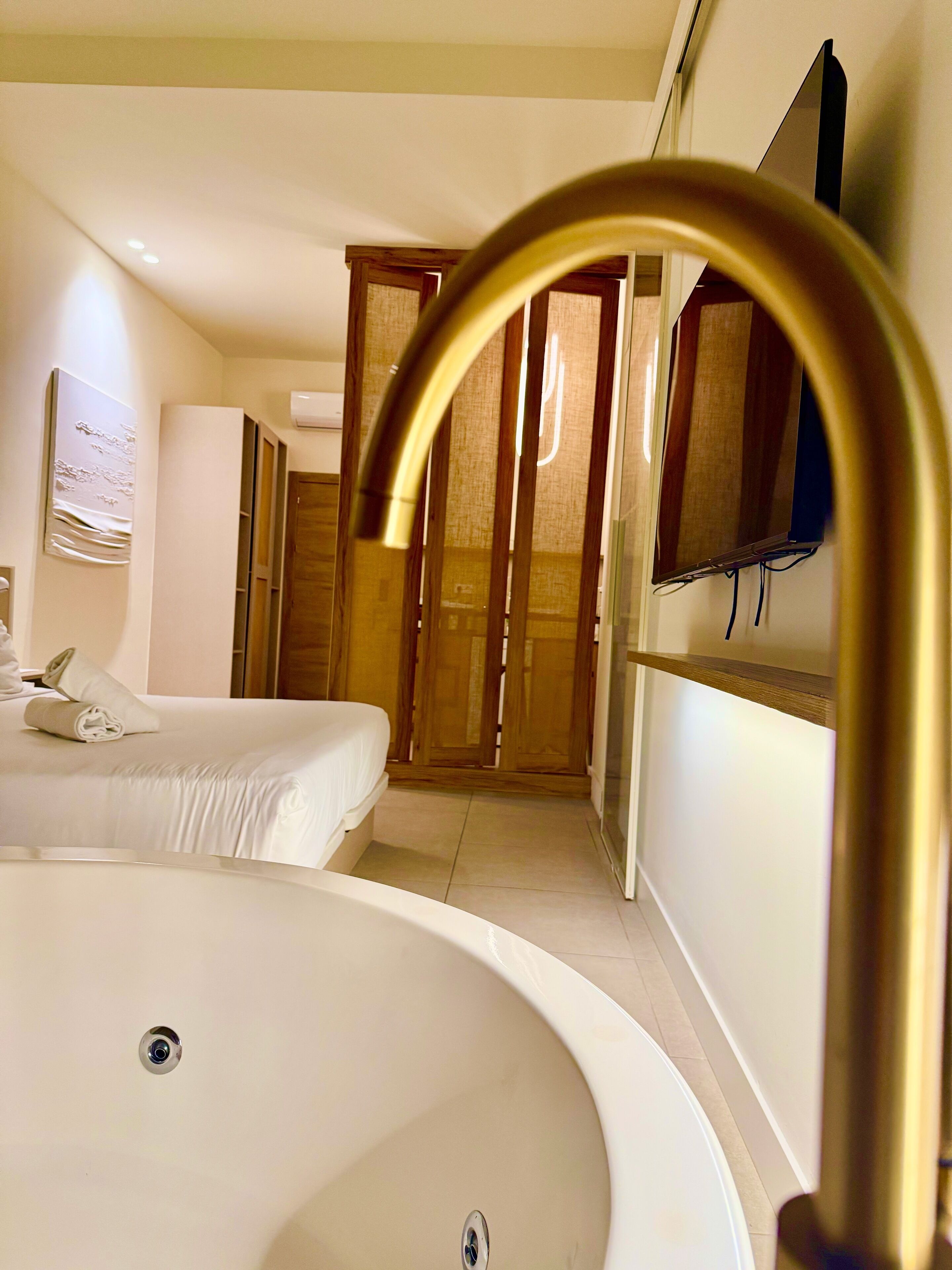 Quarto de hotel com uma banheira grande e branca, armário de madeira e uma televisão na parede.