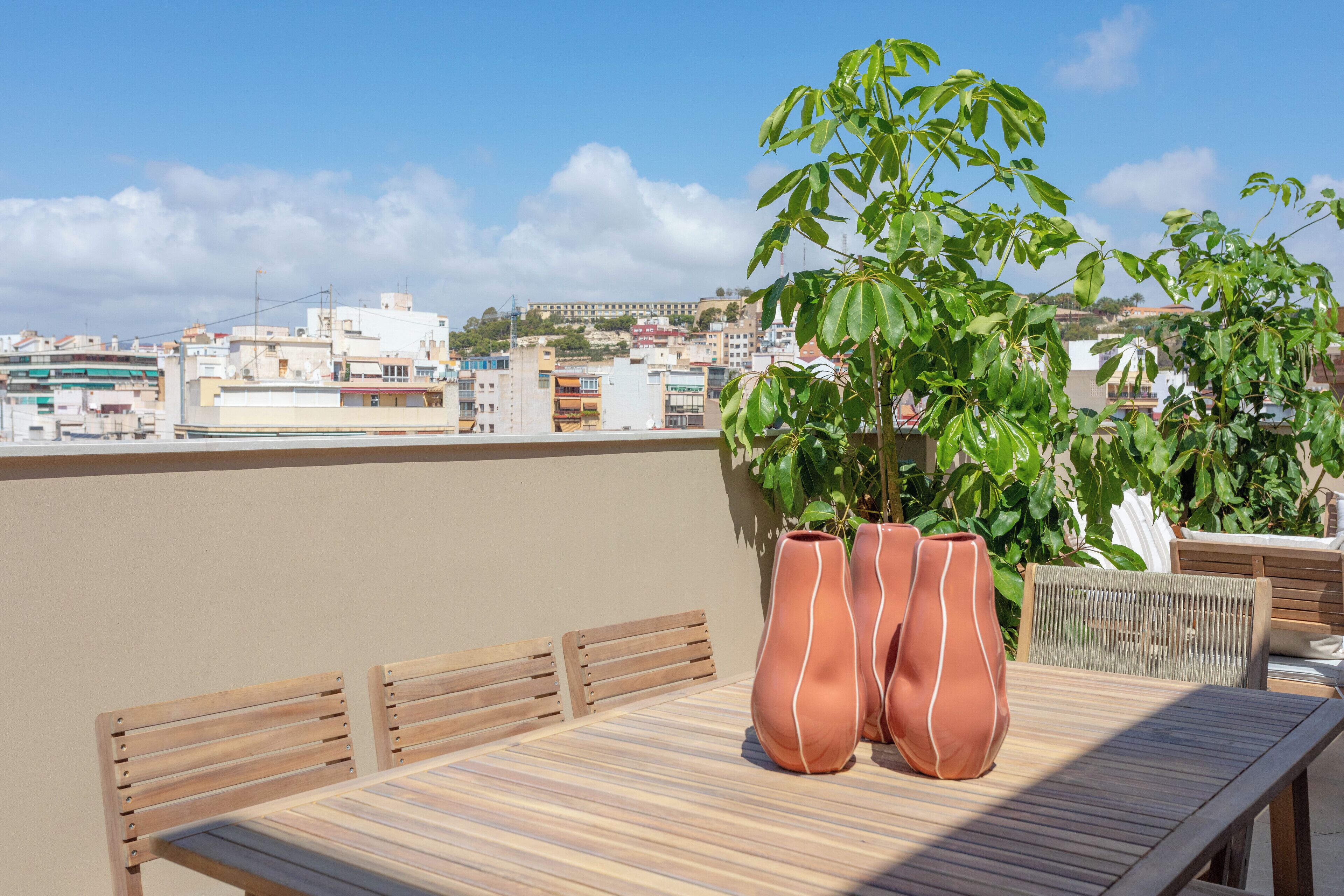 Terraço no telhado com mesa e cadeiras de madeira, vasos de terracota e vista para a paisagem urbana.