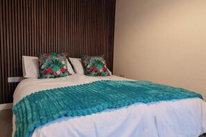 Cama com edredom branco e manta verde-água, duas almofadas com estampas florais e cabeceira de ripas de madeira.