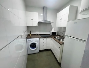 Cozinha compacta com armários brancos, máquina de lavar, micro-ondas, pia e geladeira.