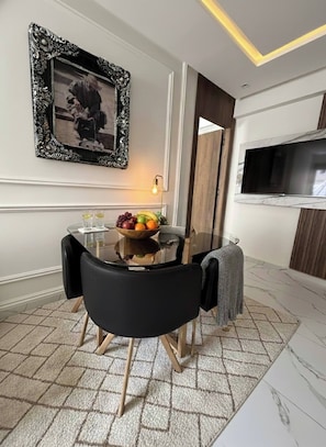 Uma sala de jantar com mesa de vidro, cadeiras pretas, uma fruteira, um quadro emoldurado e uma TV fixada na parede.