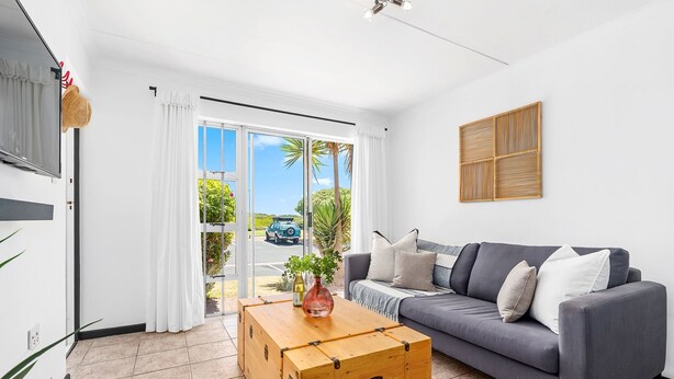 Uma sala de estar moderna com um sofá cinza, uma mesa de centro de madeira e uma grande janela com vista para palmeiras e um carro azul.