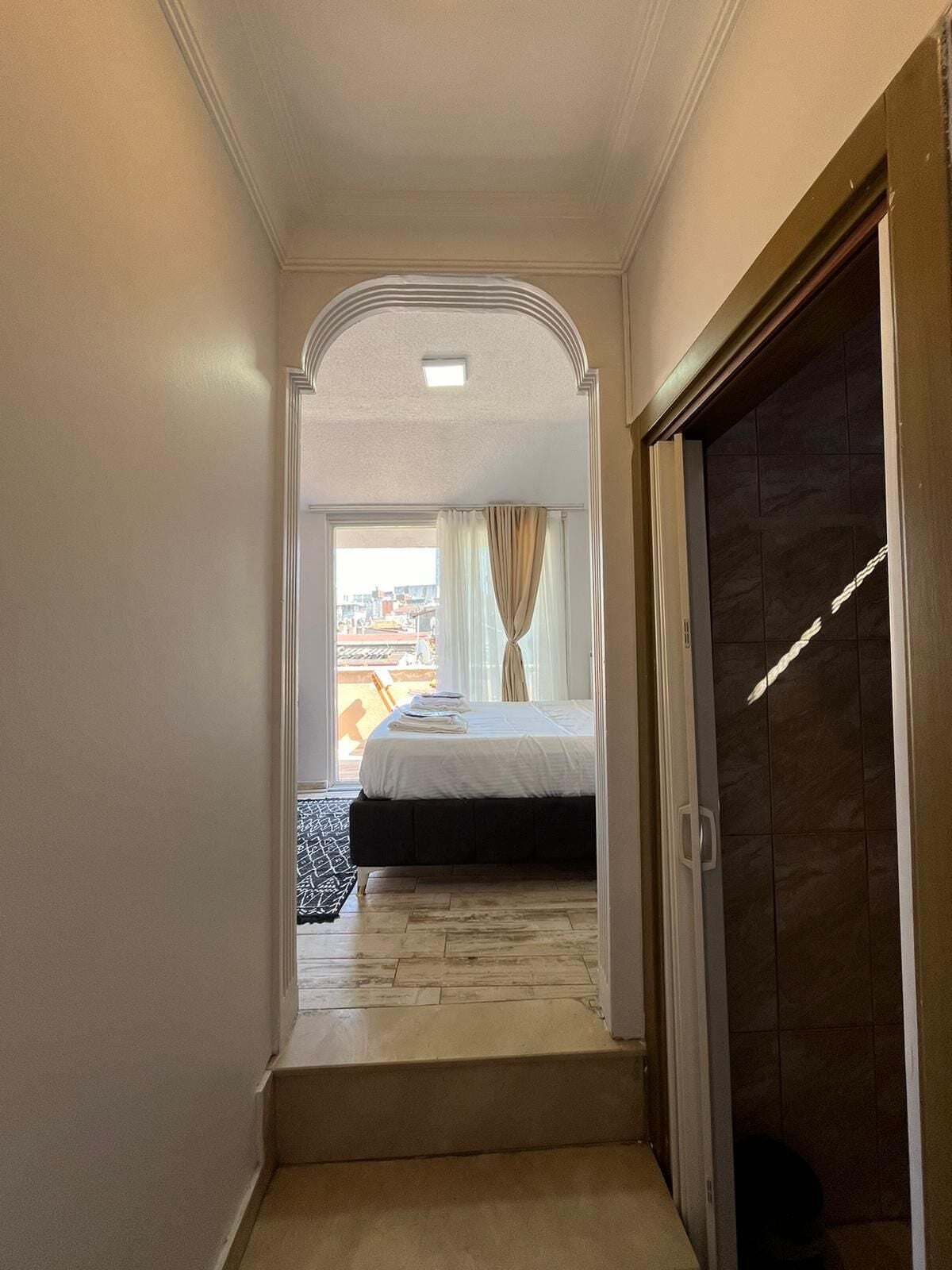 Corredor estreito que leva a um quarto com cama, vista para a cidade e porta para o banheiro.