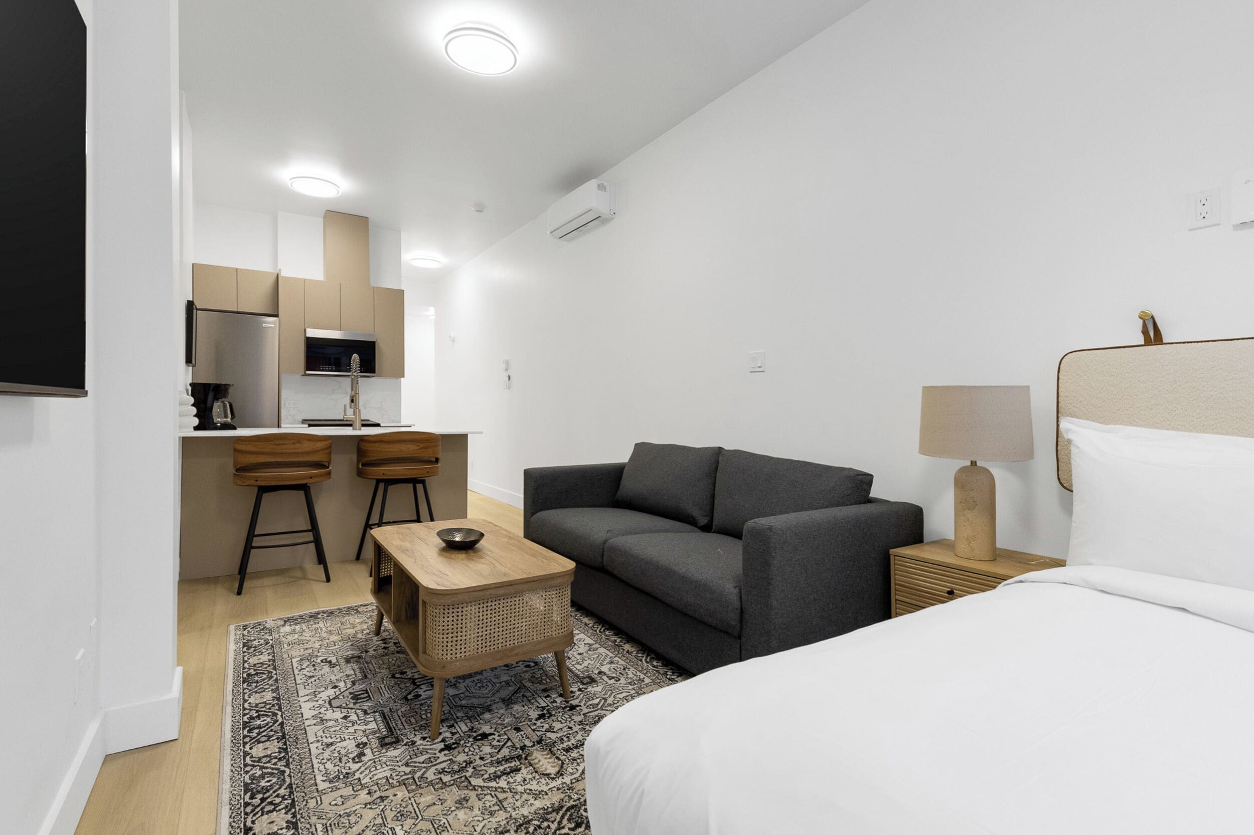 Quarto de hotel pequeno com cozinha compacta, um sofá cinza, mesa lateral de madeira, tapete estampado e uma cama com roupa de cama branca.