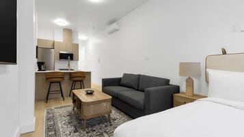 Quarto de hotel pequeno com cozinha compacta, um sofá cinza, mesa lateral de madeira, tapete estampado e uma cama com roupa de cama branca.