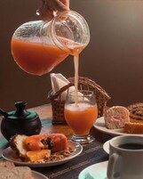 Mesa de café da manhã com jarra servindo suco de laranja em um copo, bule e diversos itens para o café.