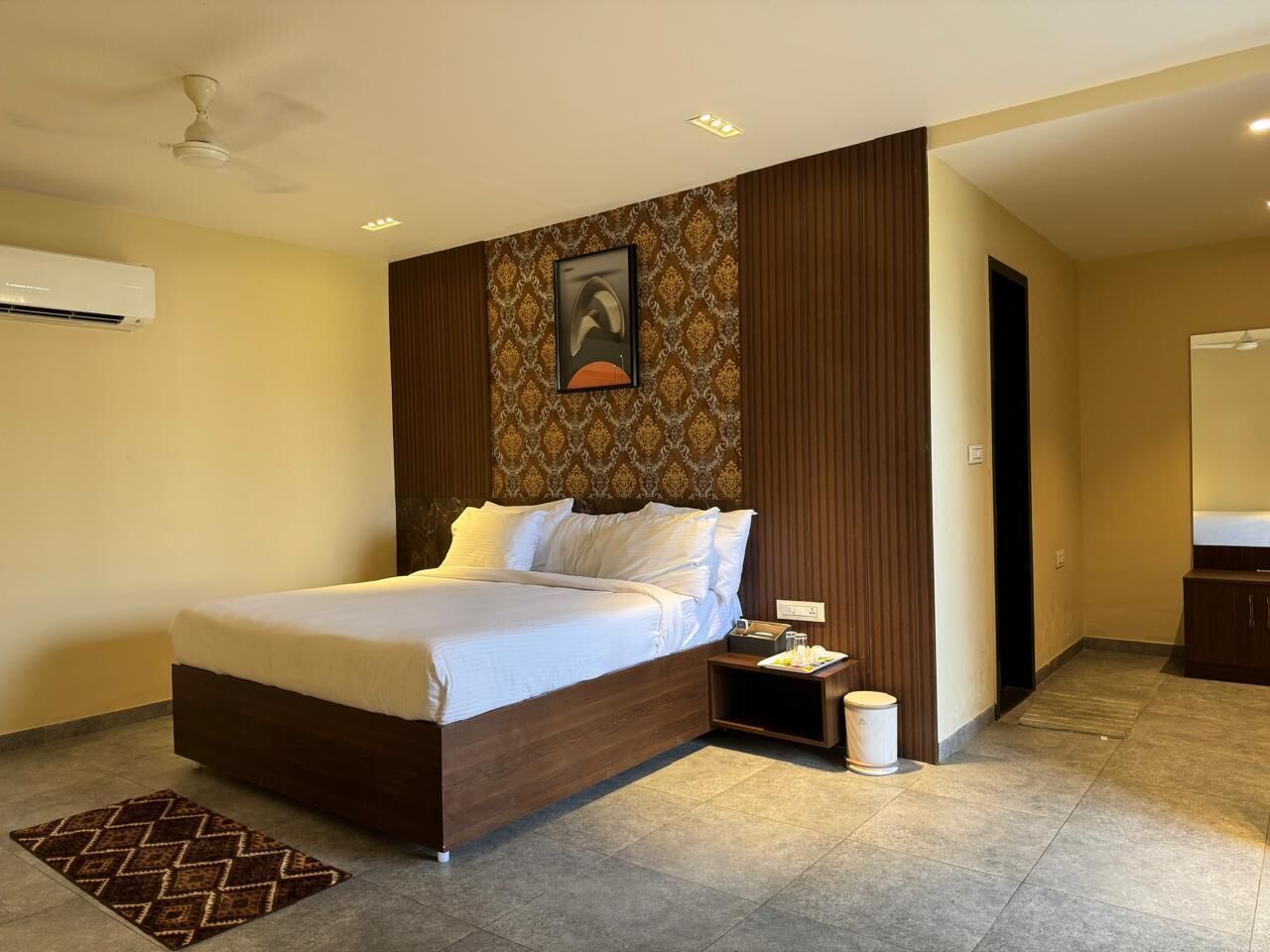Quarto de hotel moderno com uma cama grande, mesa de cabeceira, lixeira e ventilador de teto.
