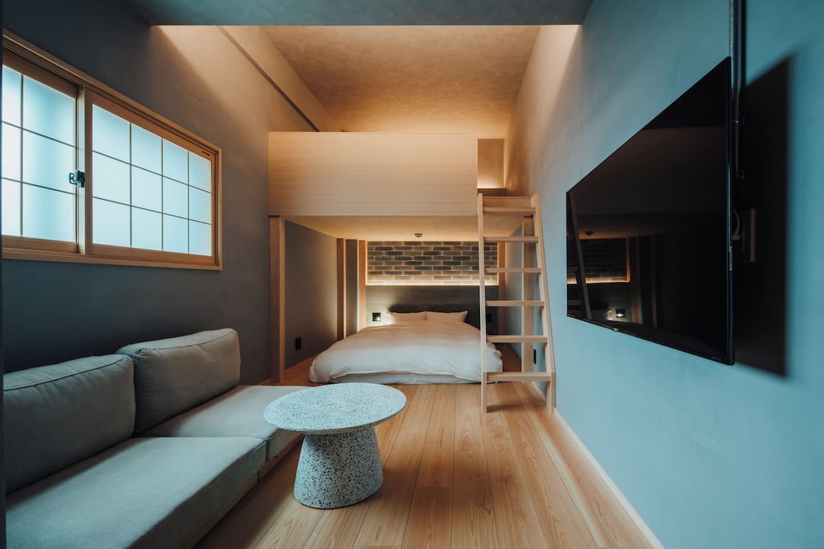 Quarto compacto com cama, escada de madeira, televisão e uma mesinha redonda.