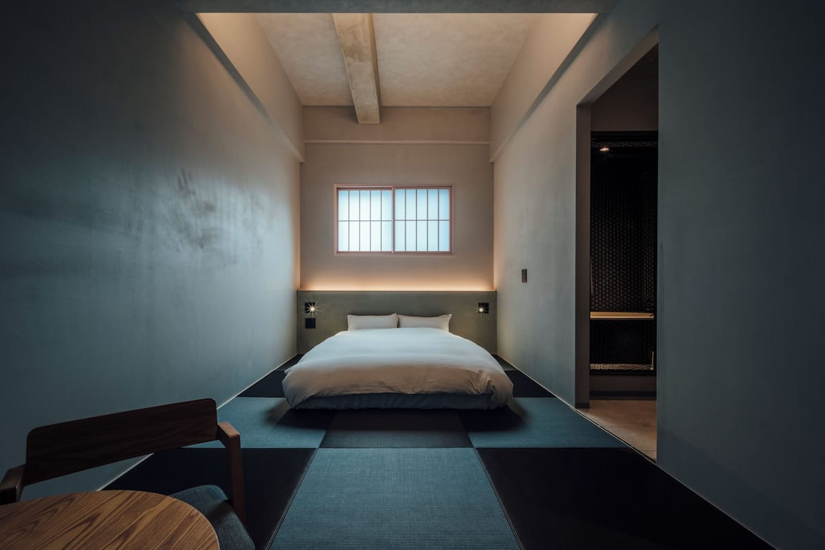 Quarto de hotel moderno com uma cama grande, uma mesa pequena e uma janela com persianas.