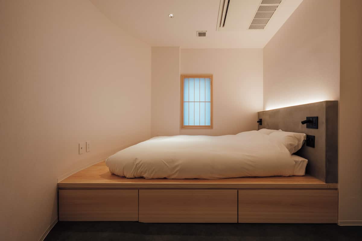 Quarto minimalista com cama embutida, uma pequena janela e luminária fixada na parede.