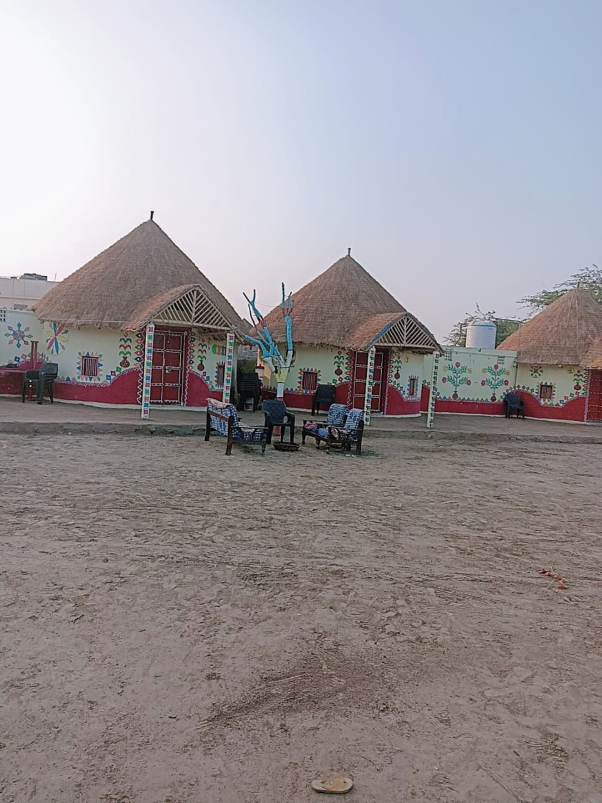 Uma fileira de cabanas com telhados de palha e fachadas coloridas, chão de areia e algumas cadeiras ao primeiro plano.