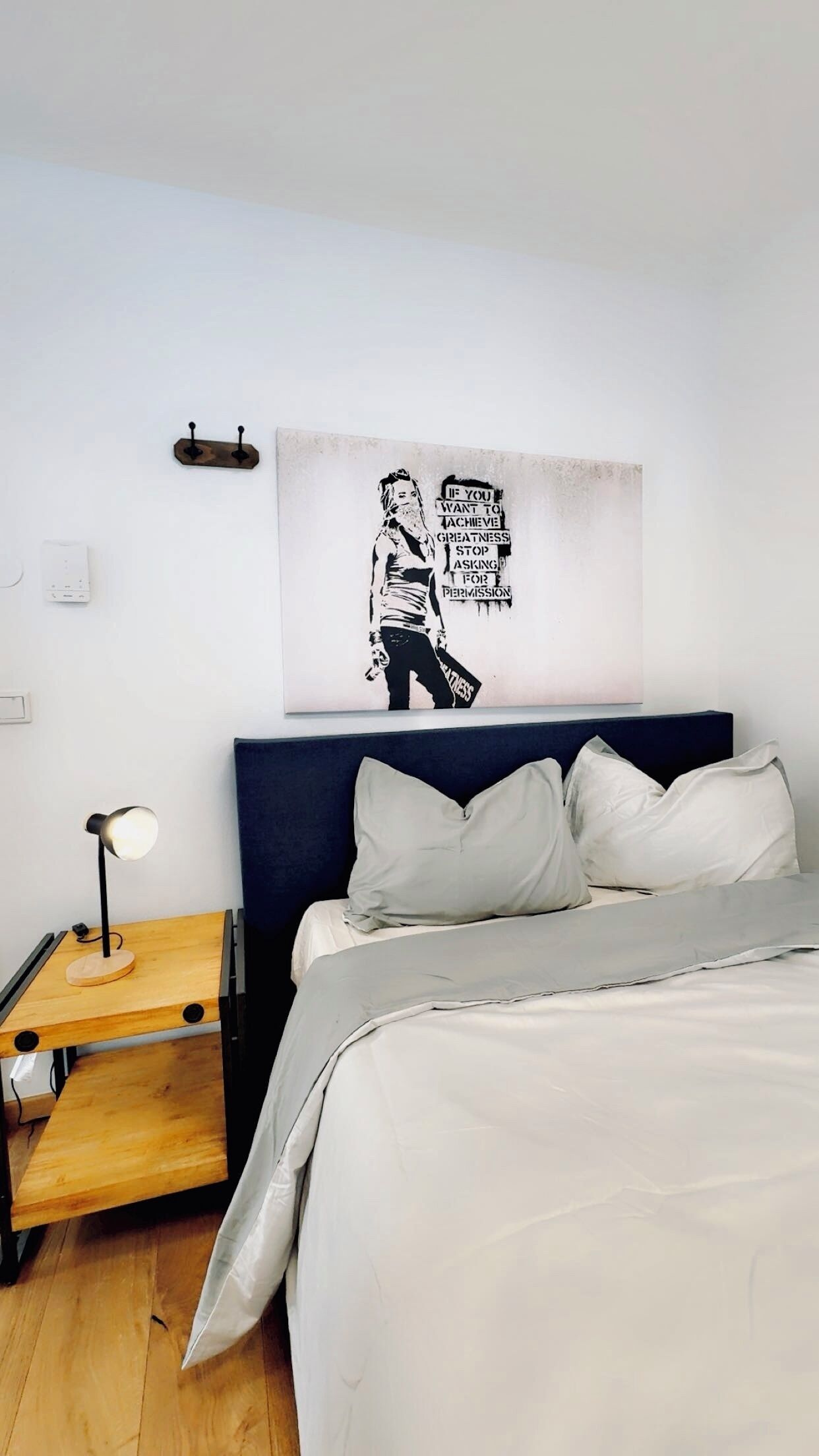 Um quarto moderno com cama, mesa de cabeceira, abajur e um pôster motivacional na parede.