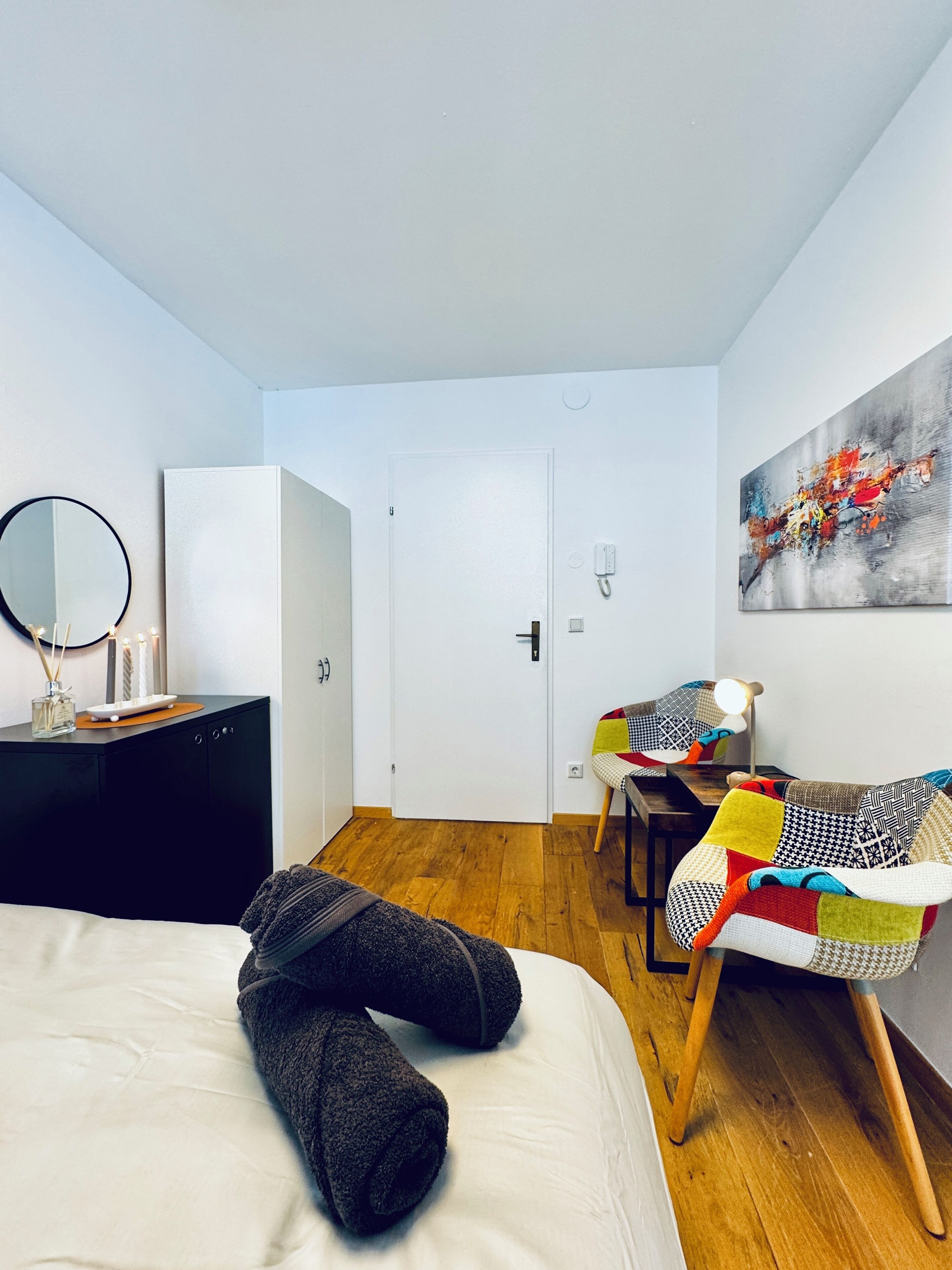 Quarto moderno com cama, mesa de cabeceira com espelho, cômoda, porta e uma cadeira colorida.
