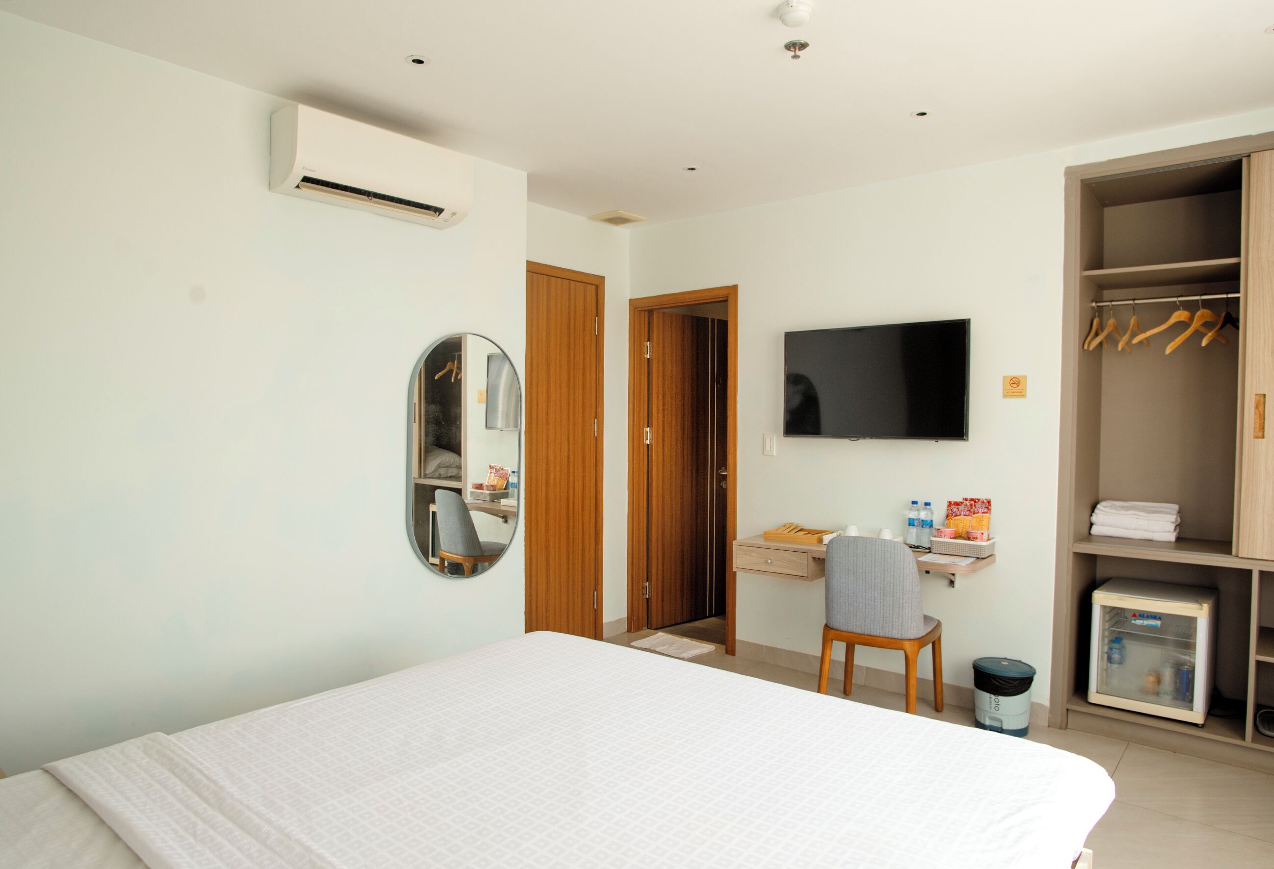 Quarto de hotel com cama, escrivaninha, cadeira, televisão, guarda-roupa e um pequeno frigorífico.