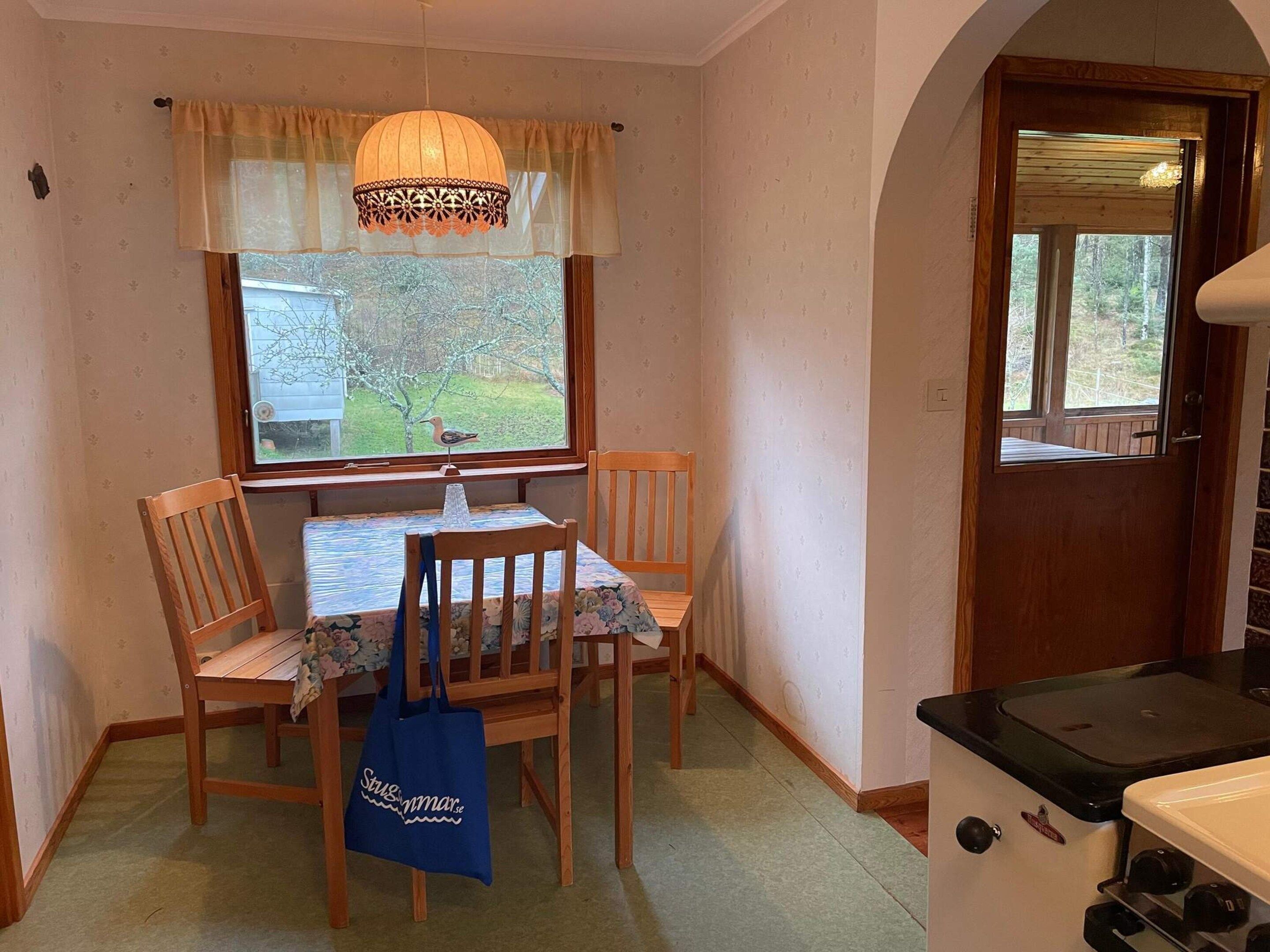 Uma área de refeições com mesa e cadeiras, uma janela com vista para árvores, um lustre e uma cozinha com fogão.