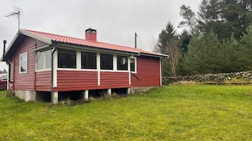Uma casa de madeira vermelha com telhado inclinado, chaminé e um muro de pedra ao fundo.