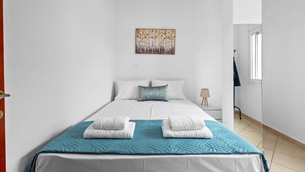 Quarto de hotel moderno com uma cama grande, roupa de cama branca, manta azul-esverdeada e um quadro na parede.