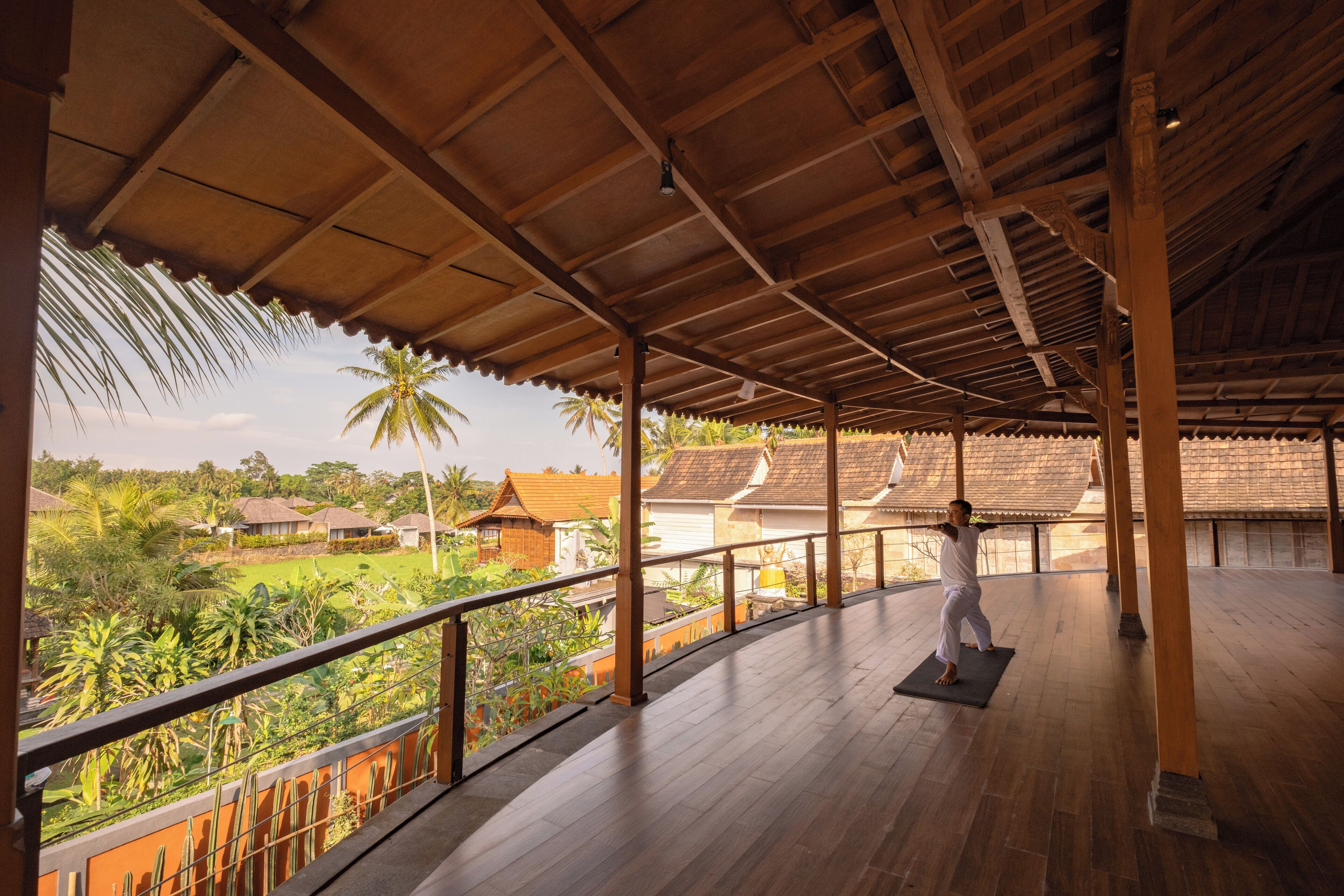 Um deck de madeira com uma pessoa praticando yoga, com vista para uma paisagem exuberante com palmeiras e edifícios tradicionais.
