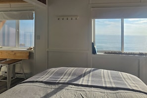 Um quarto com cama, escrivaninha e vista para o mar.
