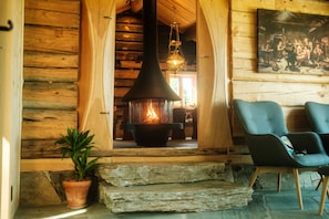 Interior de uma cabana de toras com lareira, duas poltronas estofadas e um vaso de planta.