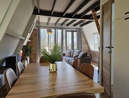 Uma sala de jantar com uma mesa de madeira comprida, cadeiras, uma planta em vaso e um sofá com almofadas.