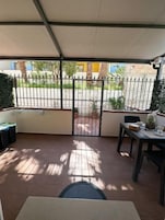 Uma área de varanda coberta com mesa e cadeiras, vista para a rua e um portão de metal.