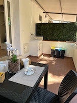 Uma varanda com mesa, cadeiras, um recipiente de vidro com pedrinhas, uma pia branca e uma parede verde com folhagem.