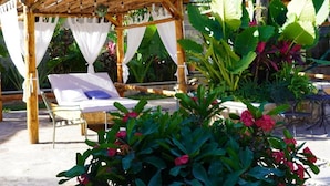 Um gazebo de madeira com teto branco, uma poltrona de descanso e plantas tropicais exuberantes.