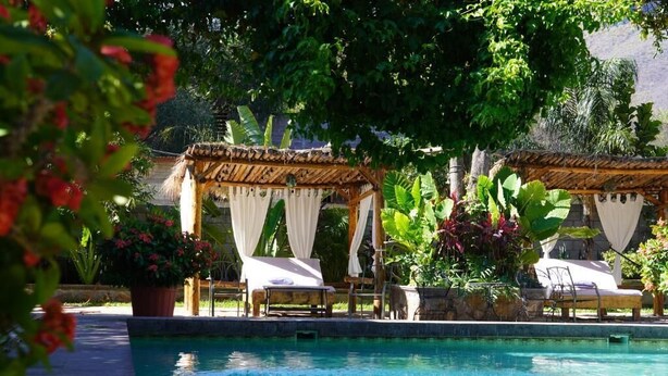 Uma área de piscina com espreguiçadeiras, uma pérgola e vegetação exuberante.